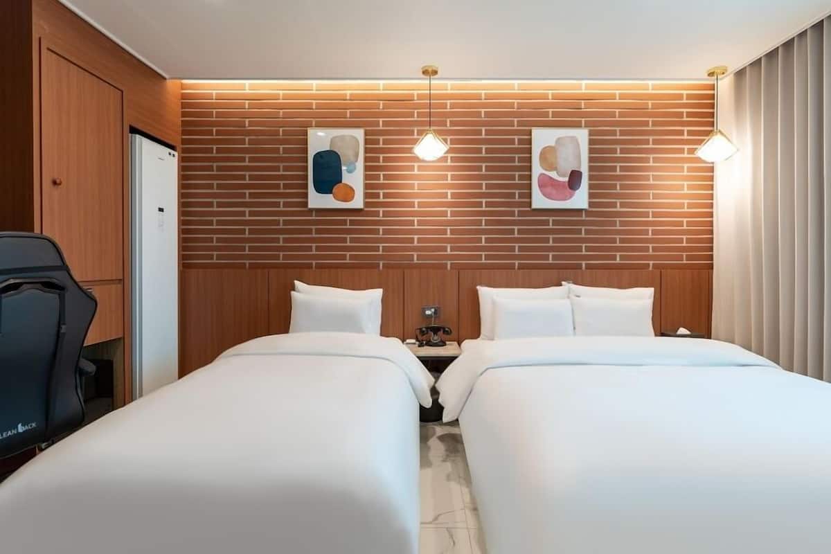 Quarto de hotel com duas camas, uma escrivaninha, uma cadeira e parede de tijolos com obras de arte.