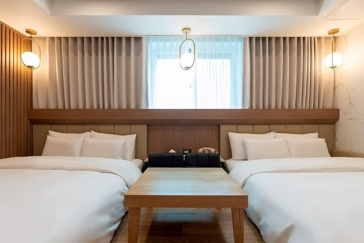 Quarto de hotel com duas camas, cabeceira de madeira, uma mesa central e duas luminárias fixadas na parede.