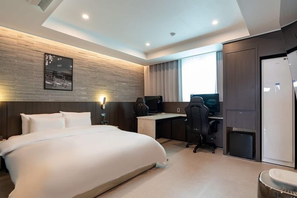 Quarto de hotel moderno com uma cama grande, uma escrivaninha com dois monitores e um pequeno espaço de estar.