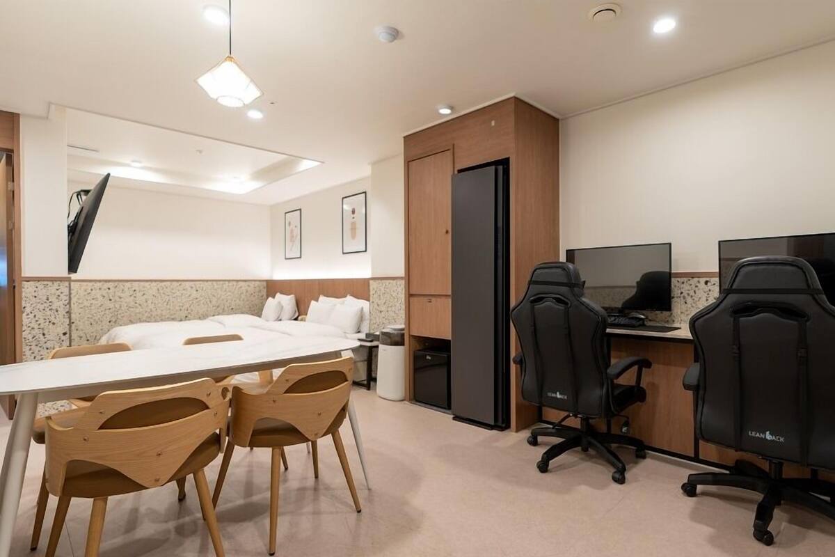 Quarto de hotel com cama, escrivaninha com dois monitores, duas cadeiras de escritório pretas, mesa de jantar de madeira com cadeiras e uma pequena lixeira.