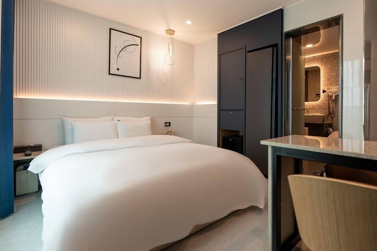 Quarto de hotel moderno com uma cama grande, uma escrivaninha com cadeira, um espelho e um banheiro visível por meio de uma divisória de vidro.
