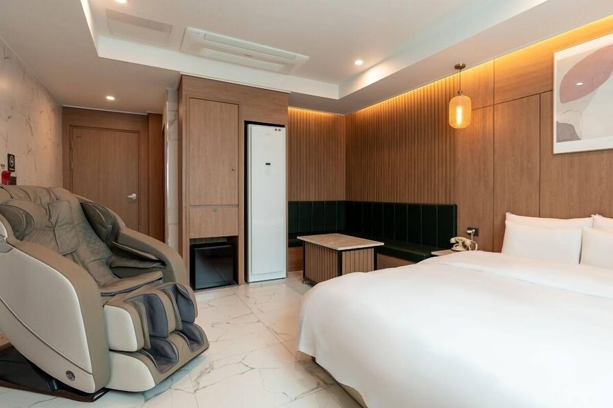 Quarto de hotel moderno com cadeira de massagem, cama grande, mesa pequena e televisão fixada na parede.