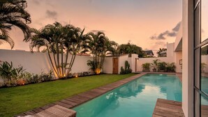Piscina com deck de madeira e espreguiçadeiras, rodeada por gramado bem cuidado e plantas tropicais.