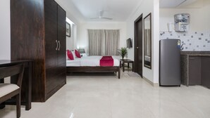 Quarto de hotel com cama, escrivaninha, guarda-roupa, ventoinha no teto, televisão e área de cozinha com frigorífico e pia.