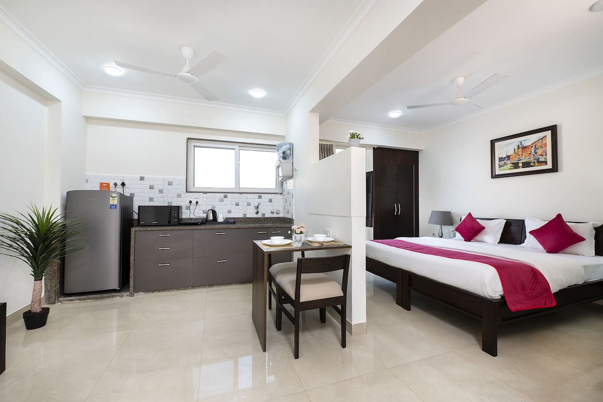 Quarto de hotel compacto com cozinha compacta, uma cama com travesseiros vermelhos, área de jantar com mesa e cadeiras e ventilador de teto.