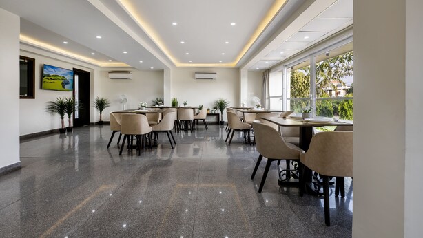 Uma sala de conferências ampla e bem iluminada, com piso lustroso, várias mesas com cadeiras e grandes janelas com vista para o exterior arborizado.