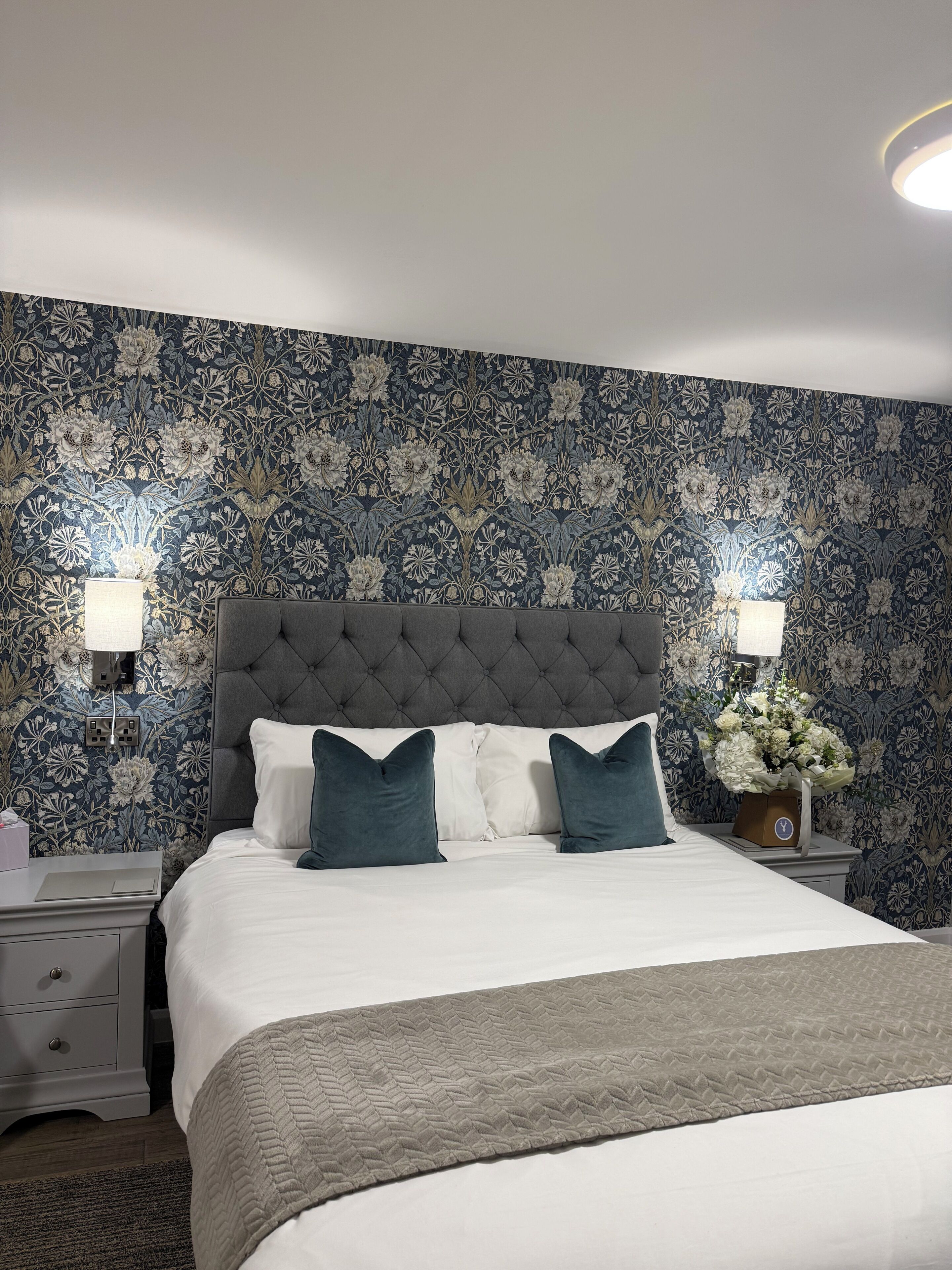 Cama bem arrumada com cabeceira cinza, duas luminárias de apoio e papel de parede com padrão floral.