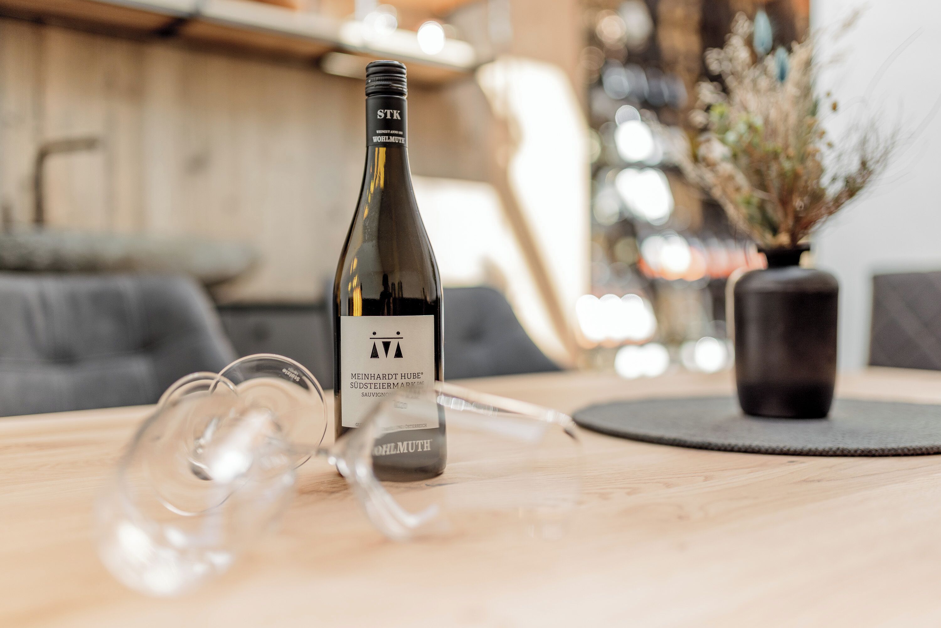 Uma garrafa de vinho, um par de óculos e um vaso com flores secas sobre uma mesa de madeira em um ambiente interno moderno.