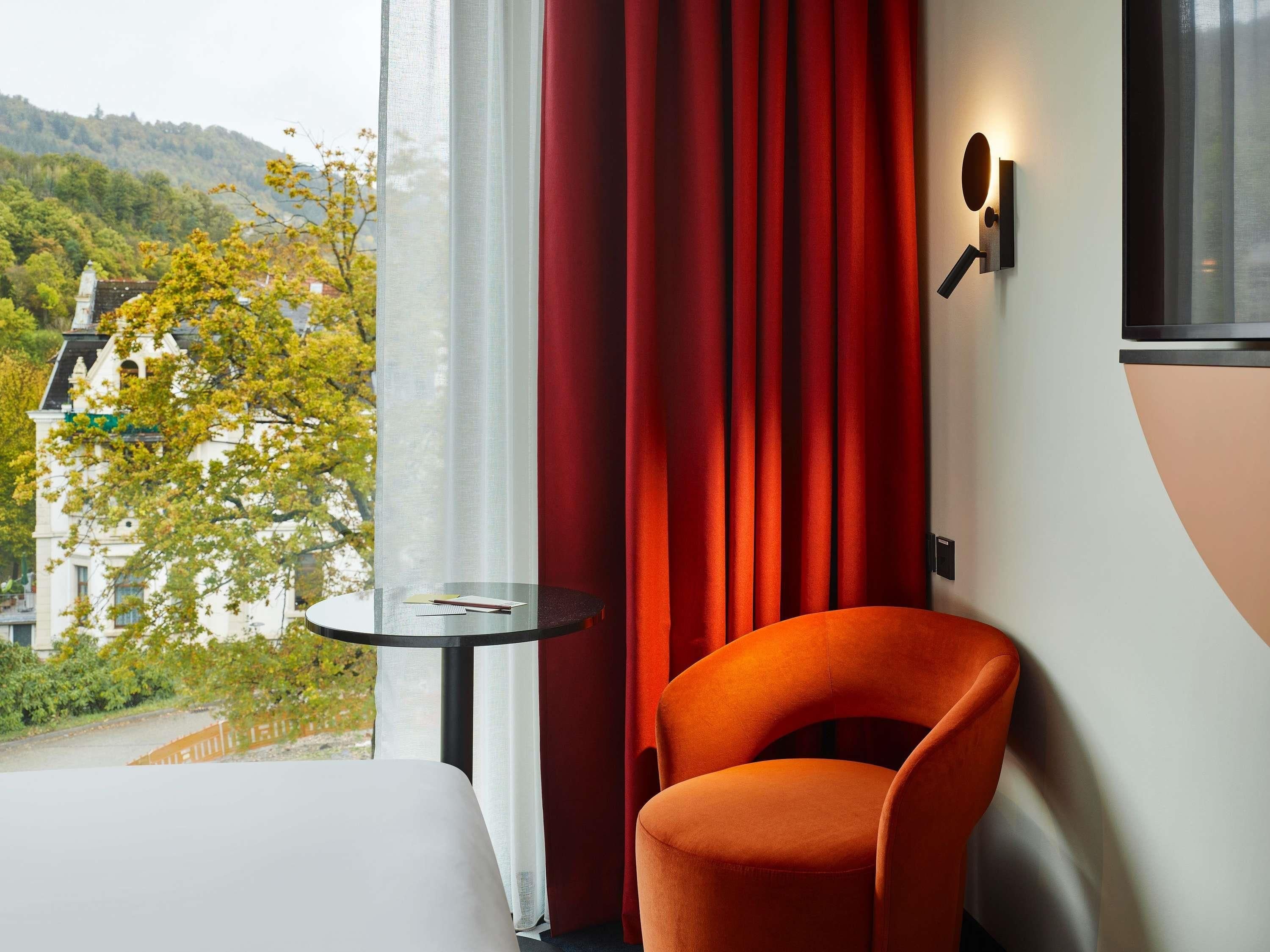 Quarto de hotel com uma cama, uma mesinha, uma cortina vermelha e vista para um edifício e árvores lá fora.