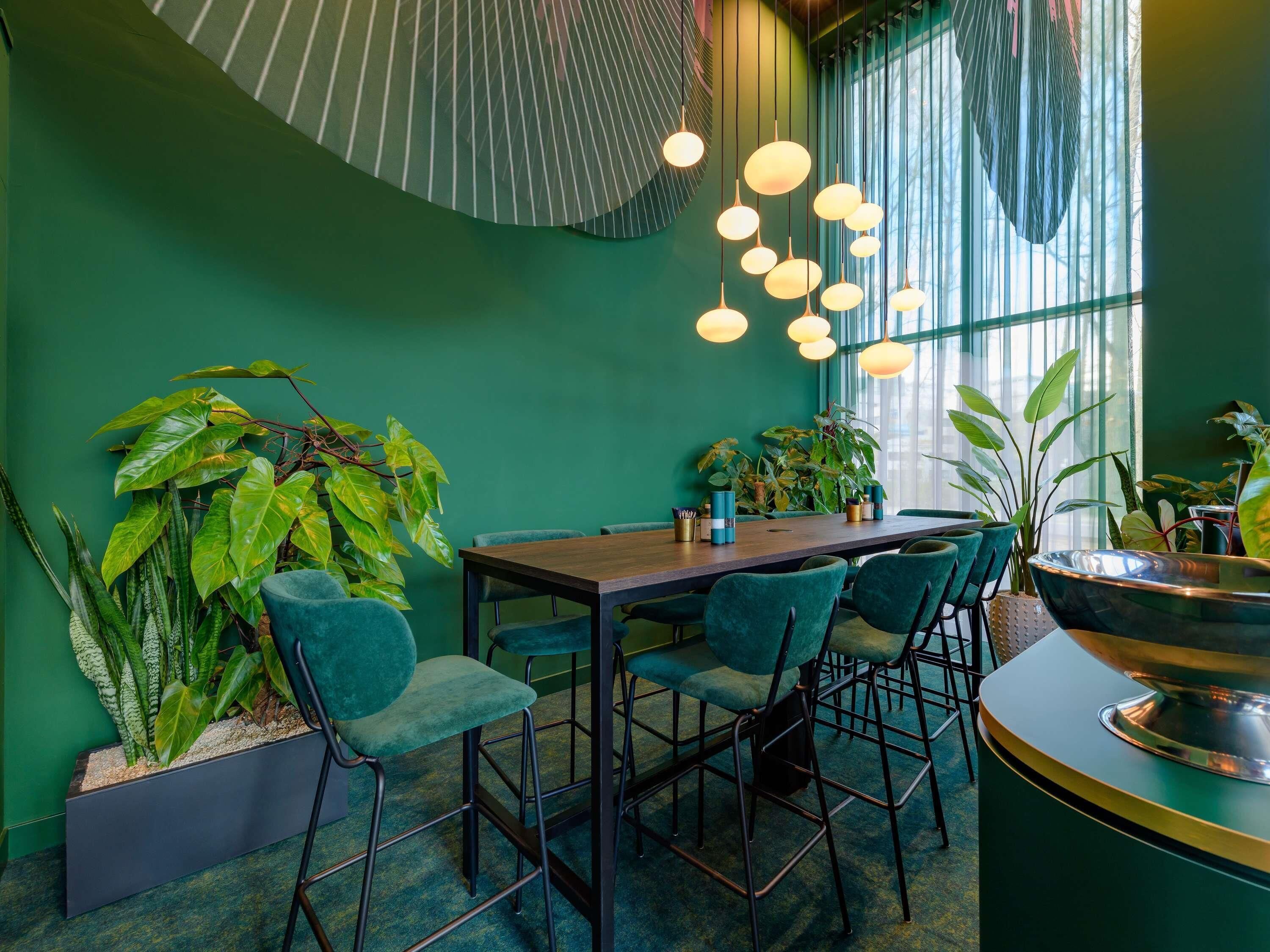 Uma sala de conferências com uma mesa comprida, cadeiras verdes e várias plantas em vasos.