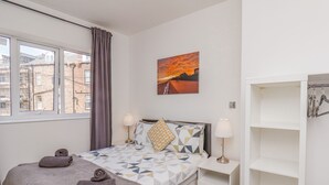 Quarto com cama, duas luminárias de cabeceira, janela com cortinas, um quadro e um guarda-roupa branco.