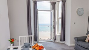 Uma área de refeições com uma mesa posta para dois, vista para a praia e um sofá cinza ao fundo.