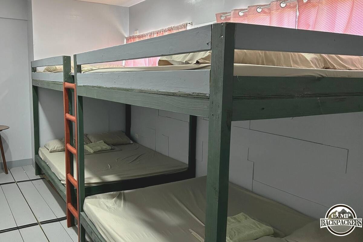 Quarto com beliche, uma mesinha e janela com cortinas.