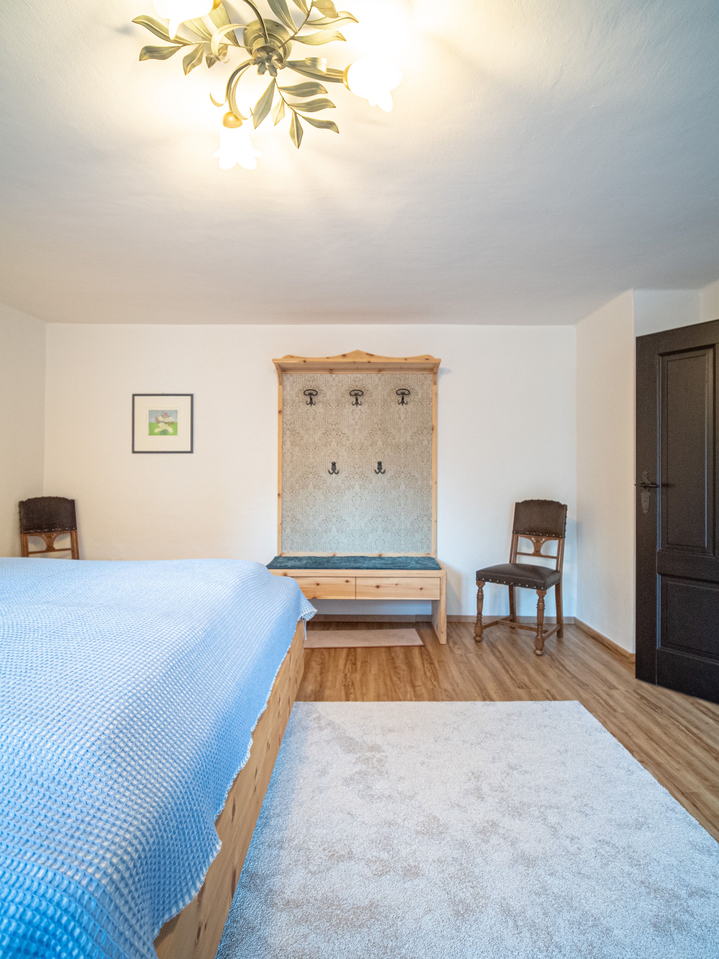 Quarto com duas camas, um banco de madeira, uma cadeira, um cabideiro e uma porta.