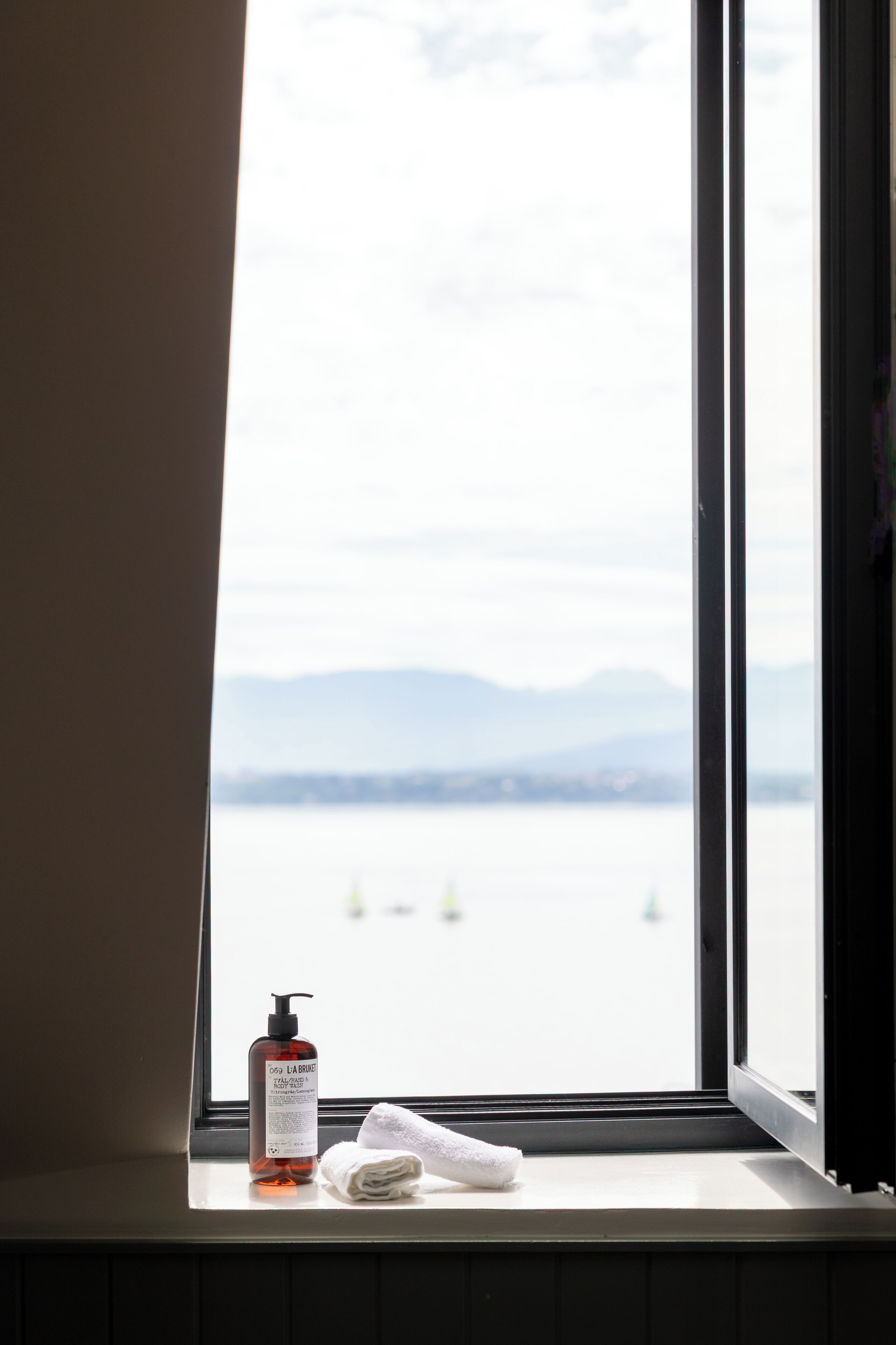 Uma embalagem de L'A Bruket de gel de banho e uma toalha branca em um parapeito de janela com vista para um corpo de água e montanhas.