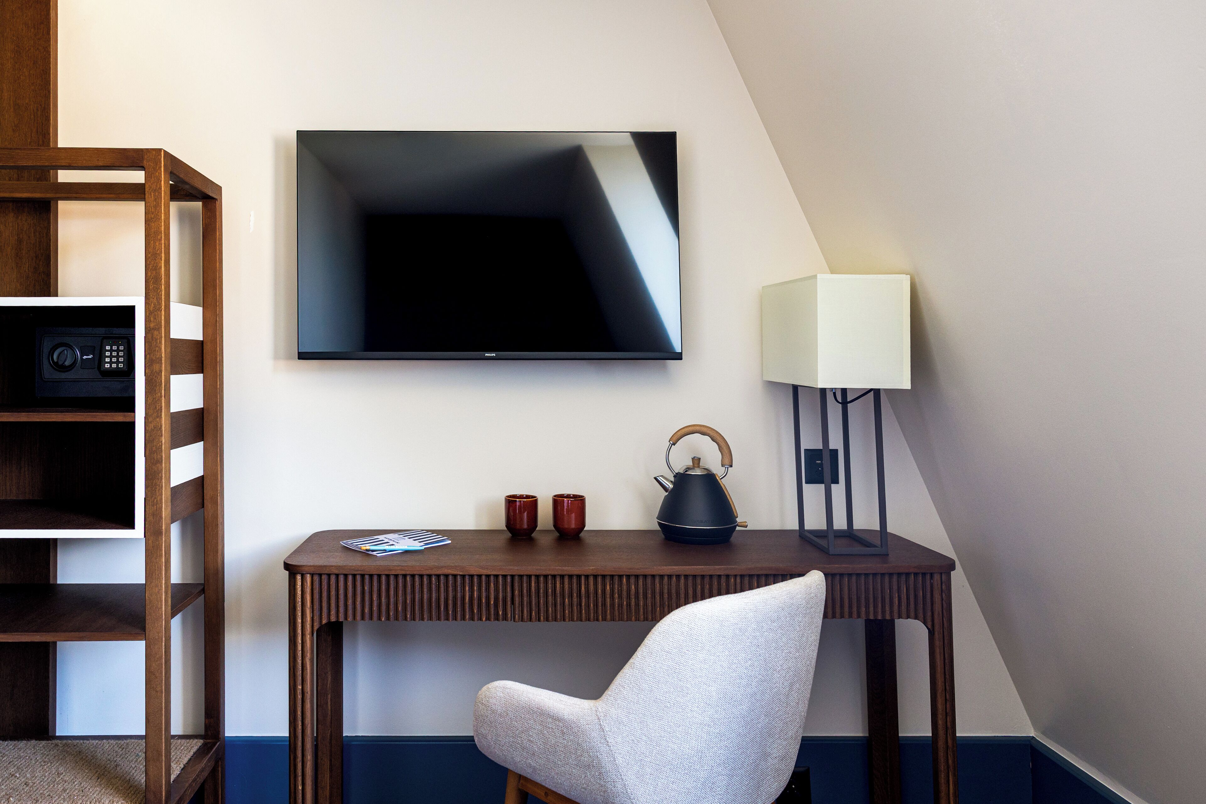 Quarto de hotel com televisão presa à parede, uma escrivaninha de madeira com uma cadeira, uma lâmpada e um bule.