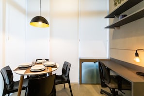 Uma área de refeições com uma mesa redonda posta para quatro pessoas, cadeiras pretas, uma luminária pendente e uma estante com uma lâmpada.