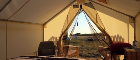 Uma tenda com cama, duas cadeiras, uma mesa e vista para um campo gramado.