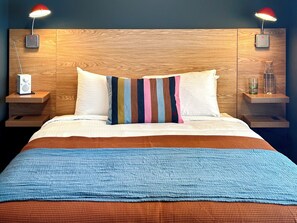 Cama bem arrumada com travesseiro listrado, cabeceira de madeira e luminárias laterais.