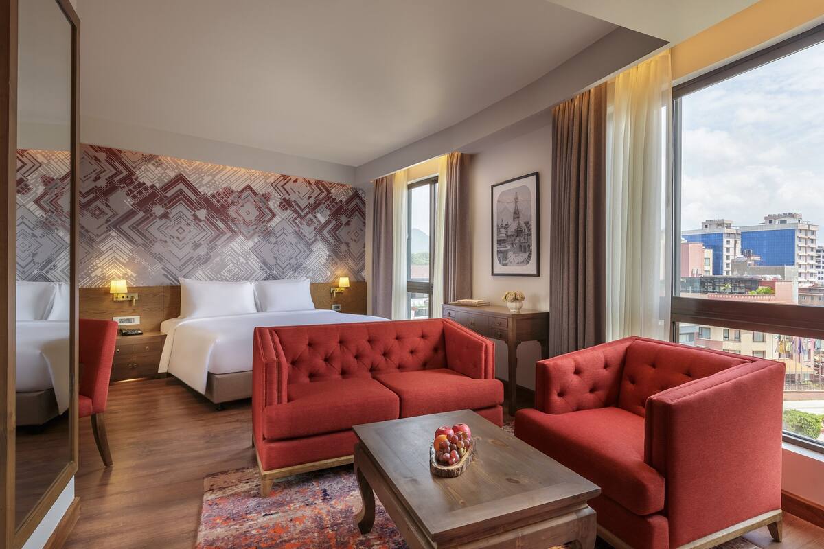 Quarto de hotel com uma cama, dois sofás vermelhos, uma mesa de centro de madeira e uma janela grande com vista para a cidade.