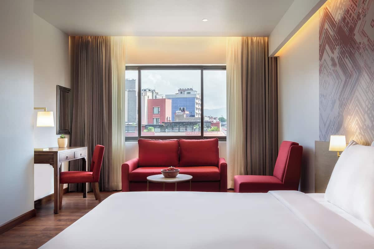 Quarto de hotel com uma cama grande, um sofá vermelho, uma escrivaninha com luminária e vista para edifícios pela janela.