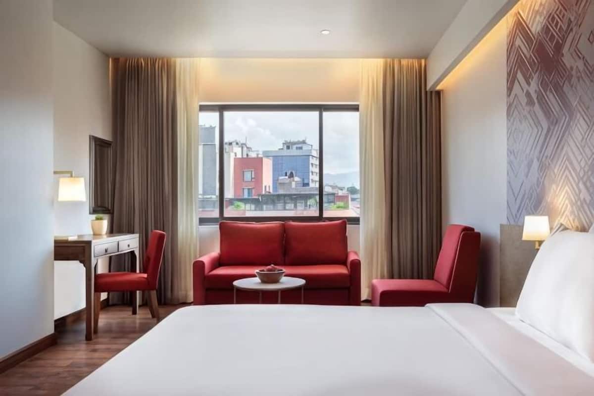 Quarto de hotel com uma cama grande, um sofá vermelho, uma pequena mesa e uma escrivaninha. Há uma janela com vista para edifícios.