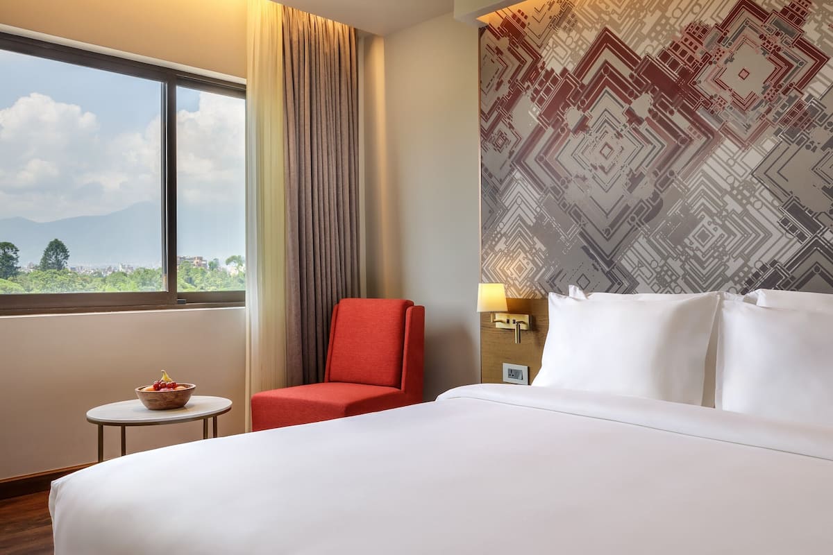 Quarto de hotel com uma cama grande, uma cadeira vermelha, uma mesa pequena com uma tigela de frutas e vista para o exterior através de uma janela.