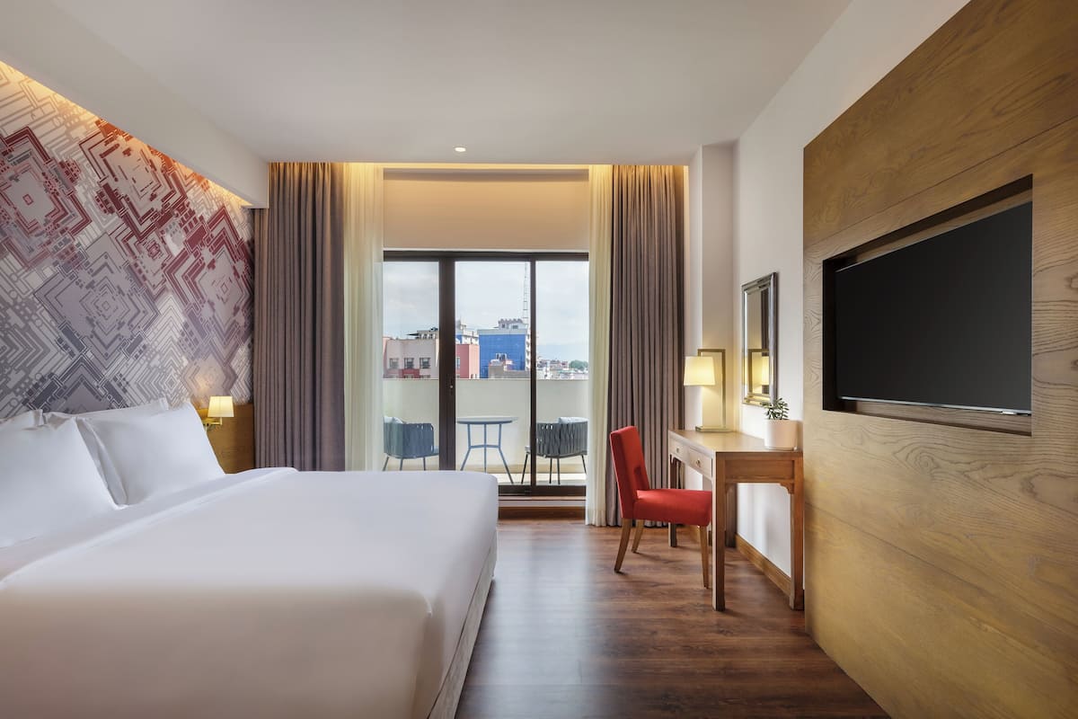 Quarto de hotel com uma cama grande, uma escrivaninha com cadeira, uma televisão e vista para a cidade pela janela.