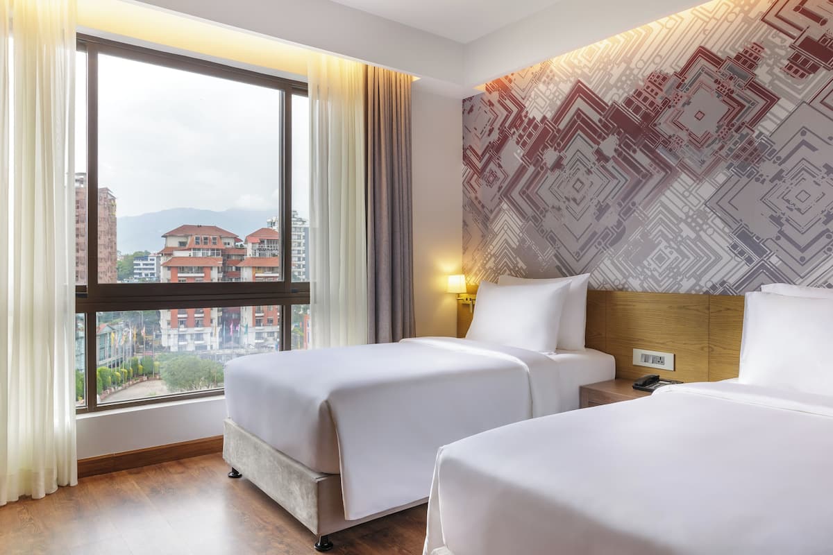 Quarto de hotel com duas camas, uma janela grande com cortinas, parede estampada e mesa de cabeceira com abajur.