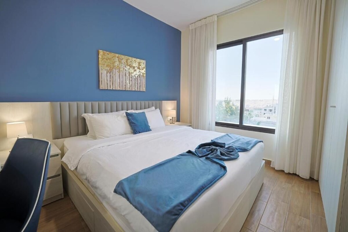 Quarto de hotel com uma cama grande, escrivaninha, cadeira, janela com cortinas e vista para a cidade.