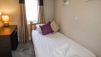 Uma cama de solteiro com roupa de cama branca e um travesseiro roxo, uma mesa de cabeceira de madeira com uma luminária e uma janela com cortinas.