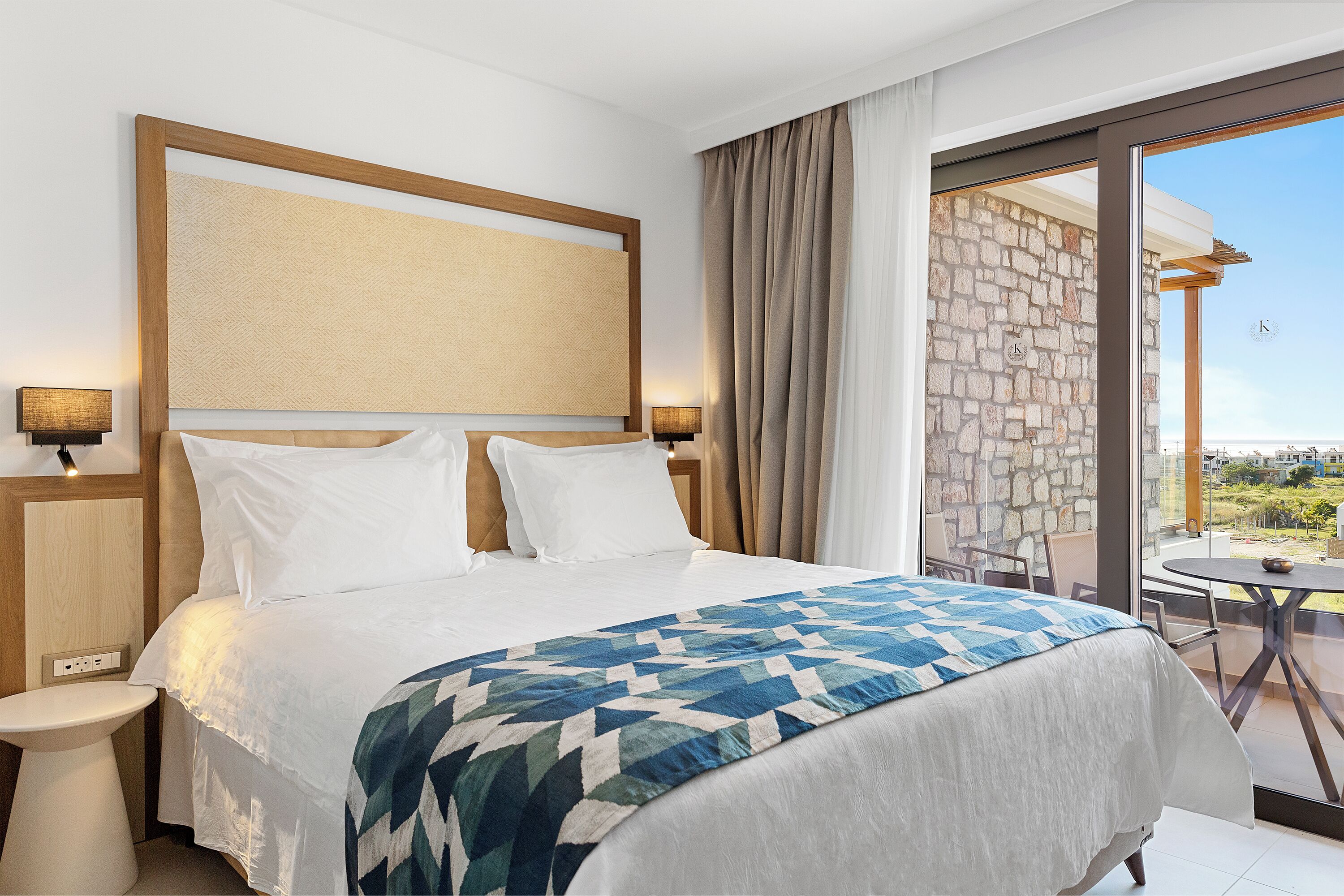 Quarto de hotel com uma cama grande, cabeceira de madeira, parede de pedra e uma varanda com mesa e cadeiras.