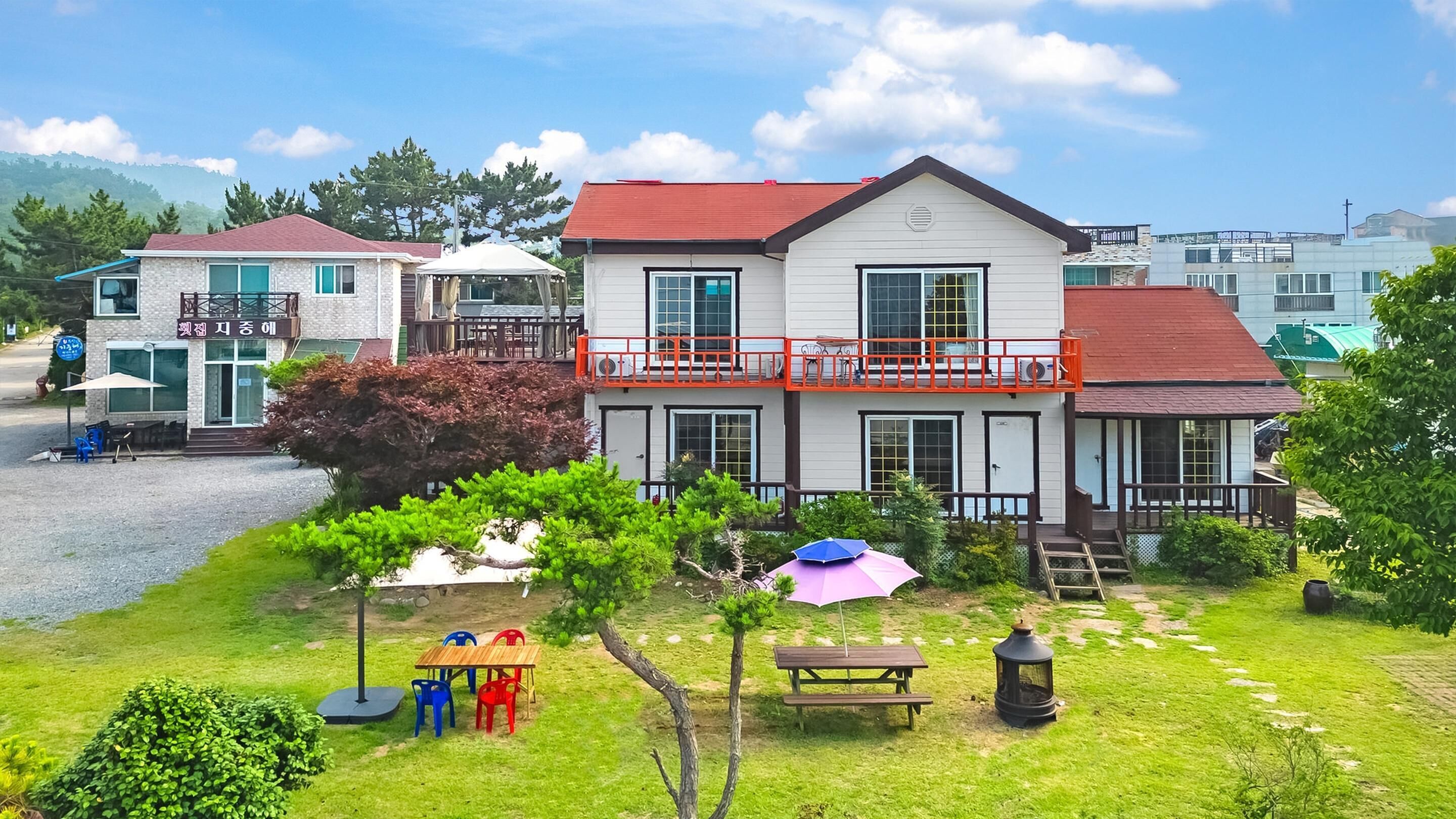 Um edifício de dois andares com varanda, uma pequena área de jardim com mesa de piquenique e uma construção com telhado vermelho ao fundo.