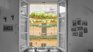 Quarto com uma janela grande que dá para um edifício amarelo com varandas. Há dois vasos pretos no parapeito da janela.