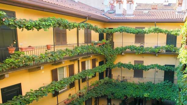 Um edifício de vários andares com paredes amarelas e telhados vermelhos, com varandas adornadas por trepadeiras e vasos de plantas.