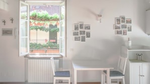 Quarto com uma mesa branca e duas cadeiras, um radiador, uma janela com vista para o verde e uma parede com quadros.
