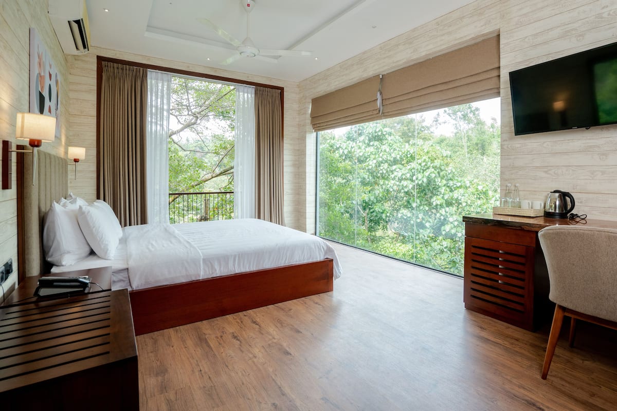 Quarto de hotel moderno com uma cama grande, uma escrivaninha com cadeira, televisão e varanda com vista para árvores.