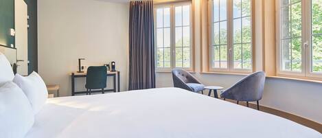 Quarto de hotel com uma cama grande, duas cadeiras, uma escrivaninha e uma janela com cortinas.
