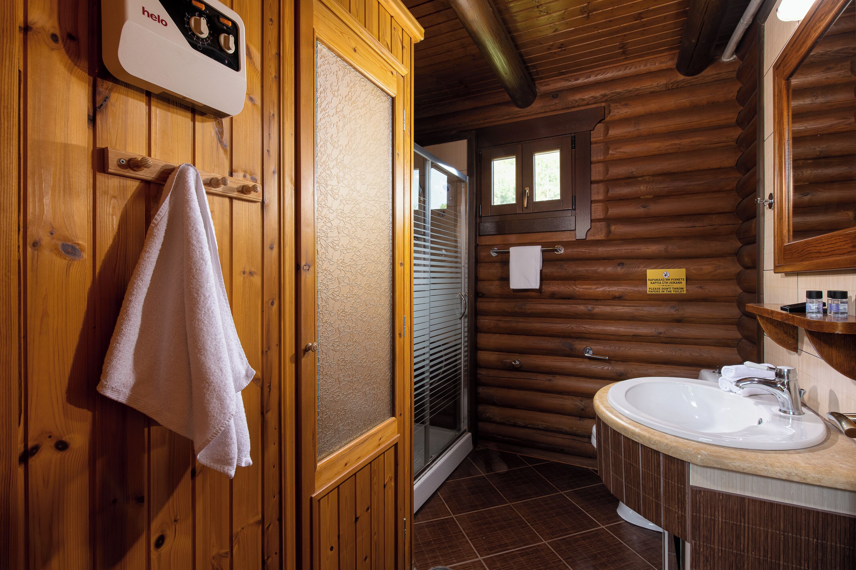 Casa de banho de cabana de madeira com porta de madeira, toalha pendurada na porta, chuveiro com porta de vidro e lavatório com espelho em cima.