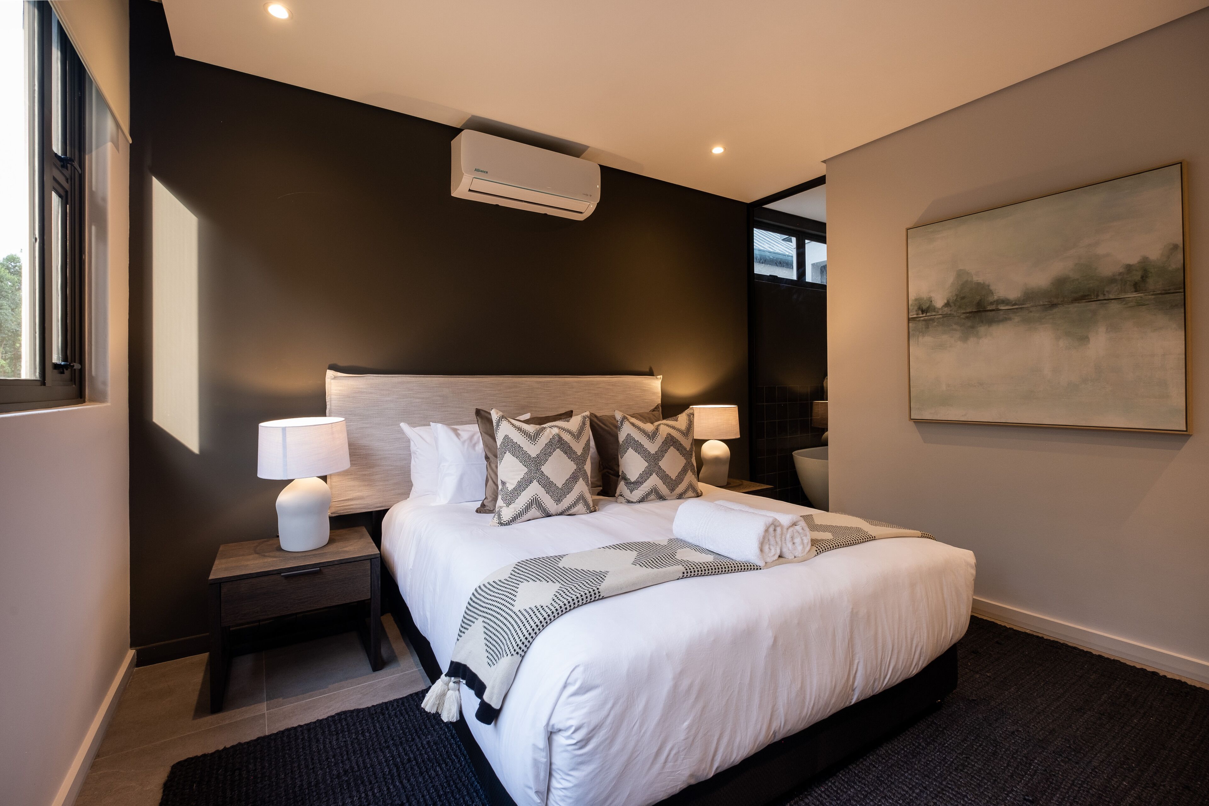 Quarto de hotel moderno com uma cama grande, uma mesa de cabeceira com luminária, ar condicionado fixado na parede e um quadro na parede.