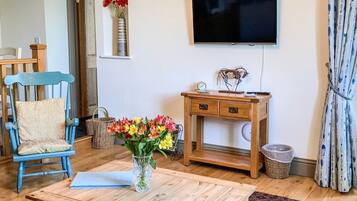 Uma sala de estar com uma cadeira de madeira, uma pequena escrivaninha, uma televisão fixada na parede e um vaso com flores sobre uma mesa.