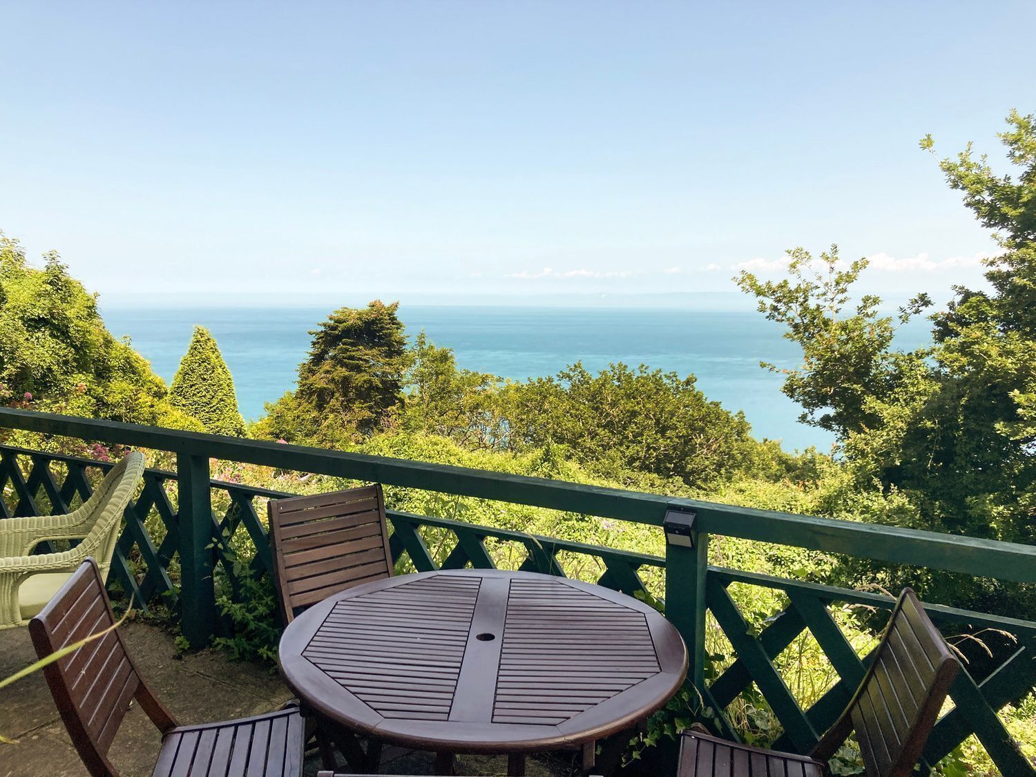 Uma varanda com uma mesa de madeira redonda e quatro cadeiras, com vista para o oceano e vegetação exuberante.