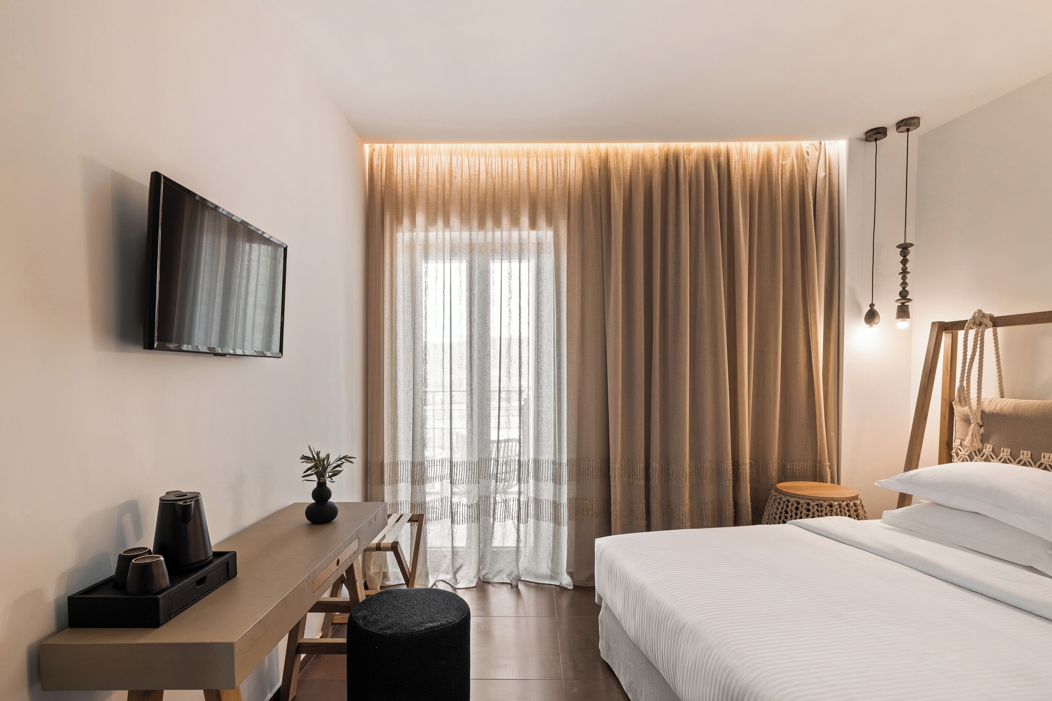 Quarto de hotel com cama, escrivaninha com cadeira, televisão presa na parede e janela com cortinas.