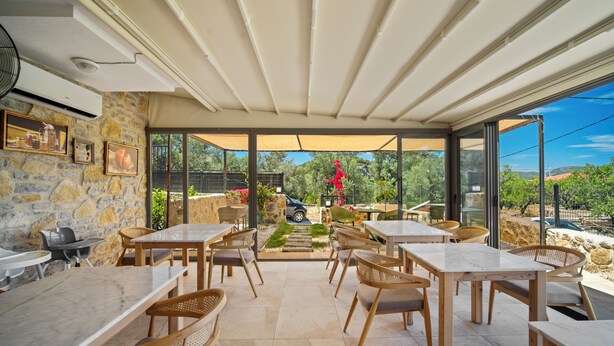 Restaurante com área externa, paredes de pedra e grandes janelas com vista para o jardim e árvores.