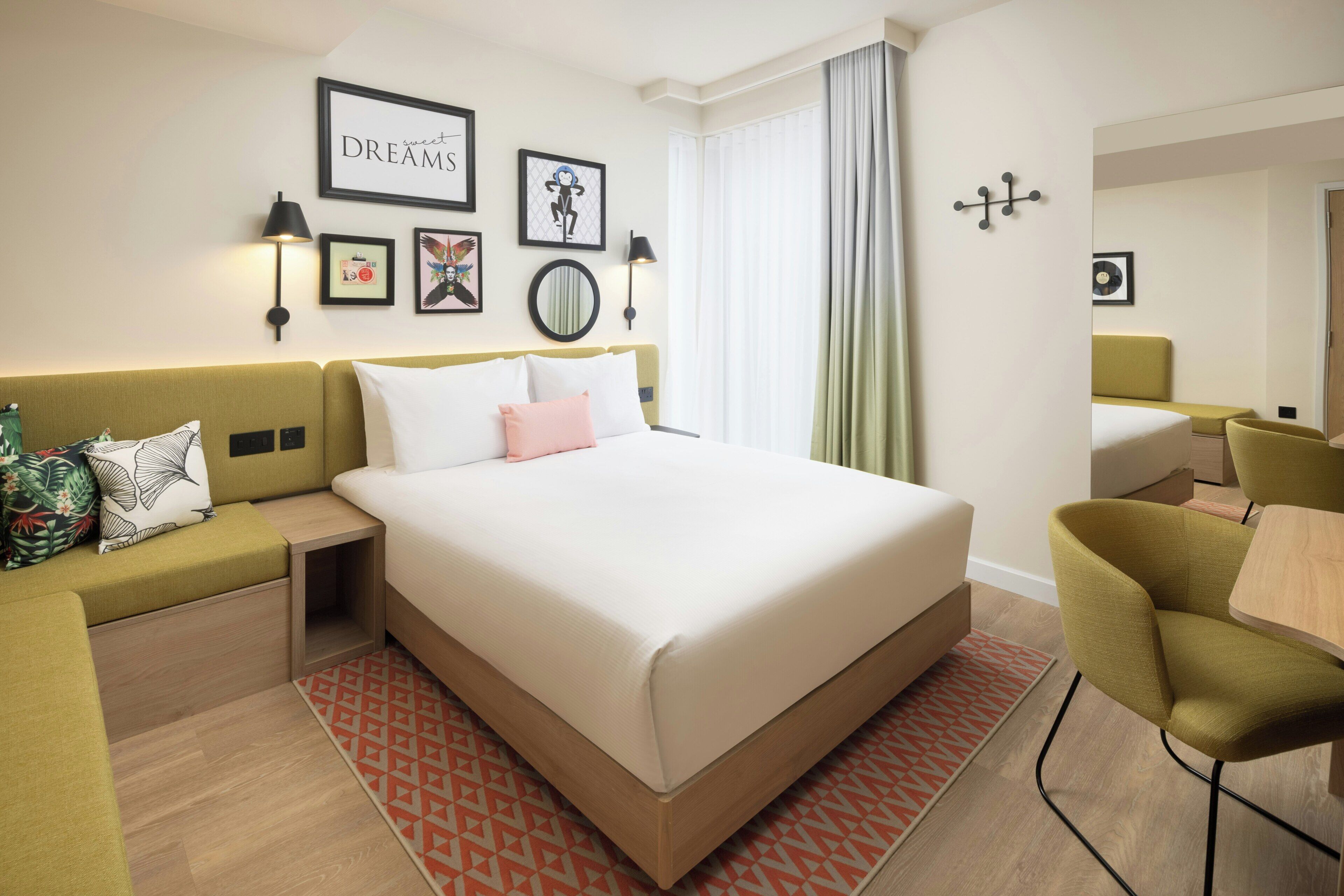 Quarto de hotel com cama, sofá, escrivaninha e cadeira. Há quadros nas paredes e uma janela com cortinas.
