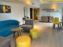 Uma sala de estar moderna com um sofá azul, uma poltrona cinza e pufes amarelos. Uma pequena mesa com petiscos está em frente ao sofá. O espaço tem um design minimalista, com um espelho grande e um armário ao fundo.