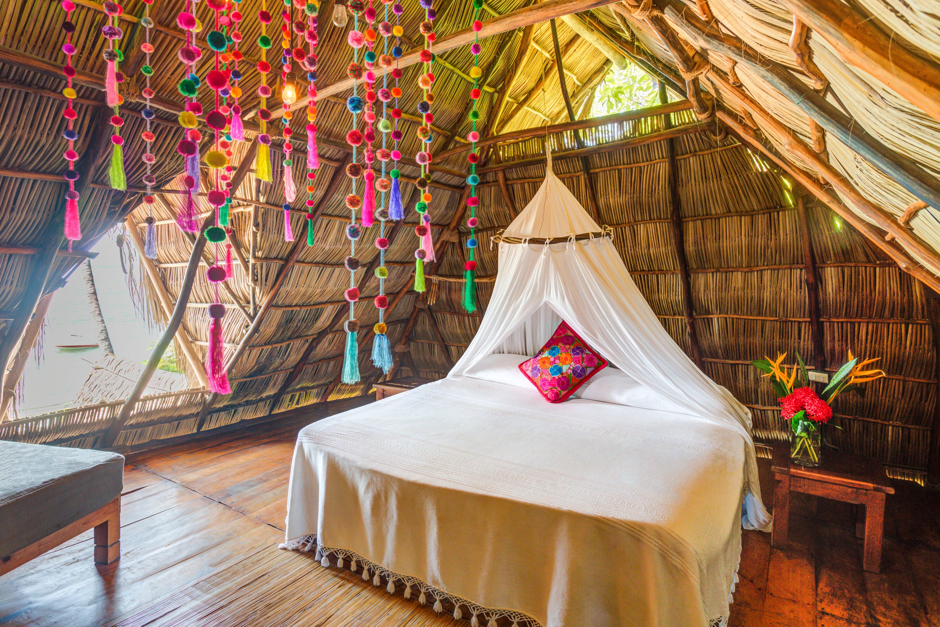 Quarto com telhado de colmo, uma cama, decorações penduradas e uma pequena mesa com flores.