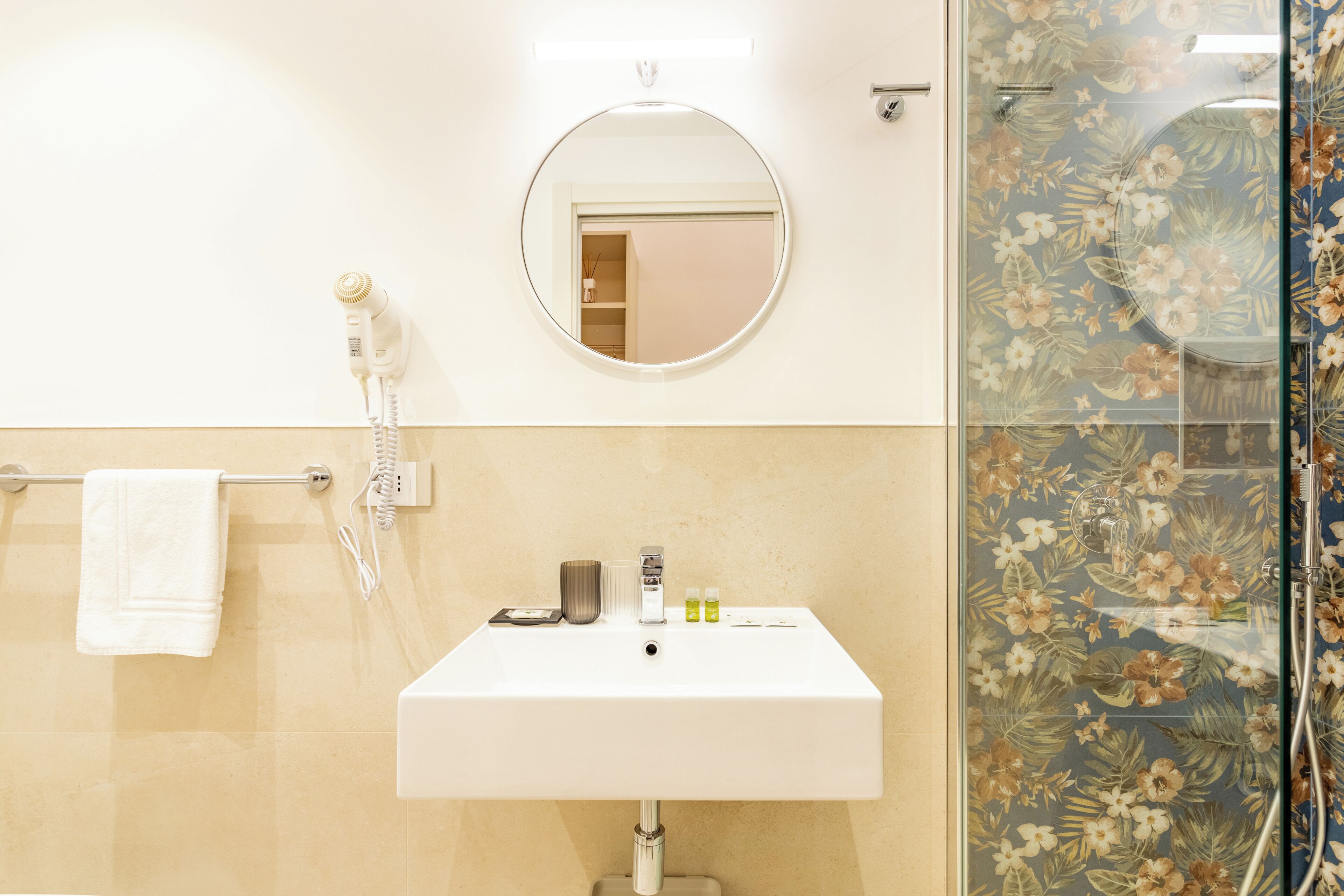 Casa de banho com lavatório branco, espelho redondo, secador de cabelo e chuveiro com papel de parede floral.