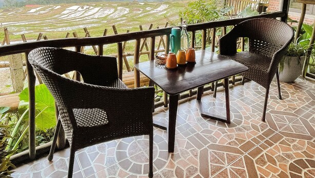 Um terraço com telhado de palha, cadeiras de vime, mesa com garrafa e copos, e vista para campos em socalcos e montanhas.