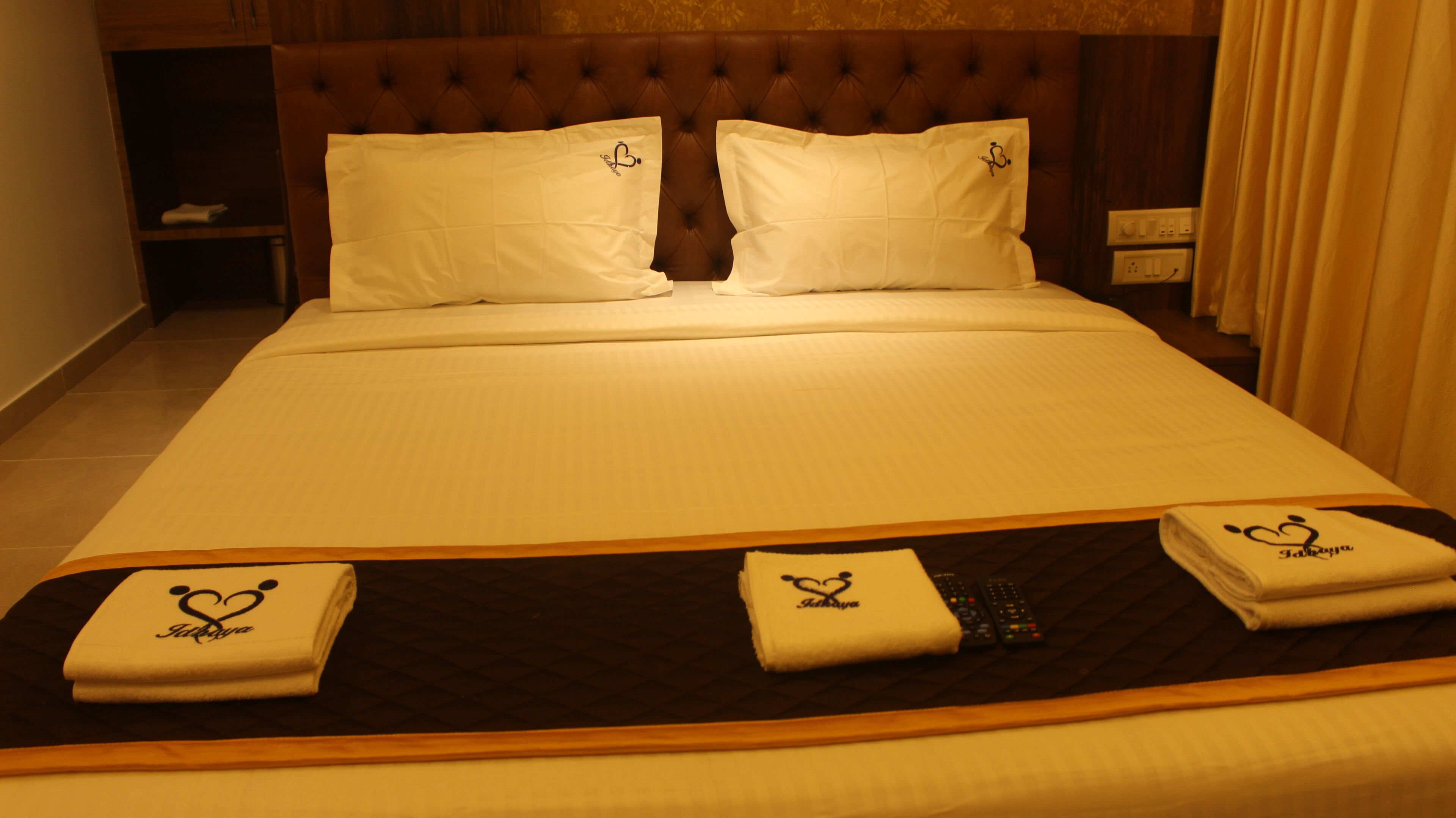 Cama bem arrumada com lençóis brancos, dois travesseiros e uma toalha dobrada com desenho de coração sobre um corredor de cama escuro.